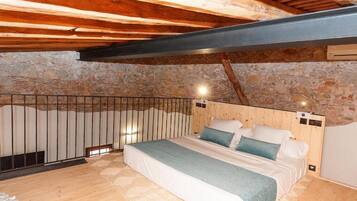 Quarto com teto de madeira, paredes de pedra, cama com colcha branca e azul, e uma pequena janela.
