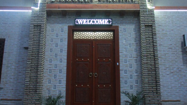 Entrada de hotel com um letreiro de neon, placa de boas-vindas e porta de madeira.
