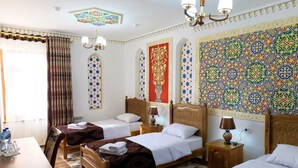 Quarto de hotel com duas camas, uma escrivaninha de madeira e uma mesa com garrafa de água e copos.
