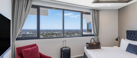 Quarto de hotel com cama, poltrona vermelha, mala e vista para a cidade através de grandes janelas.