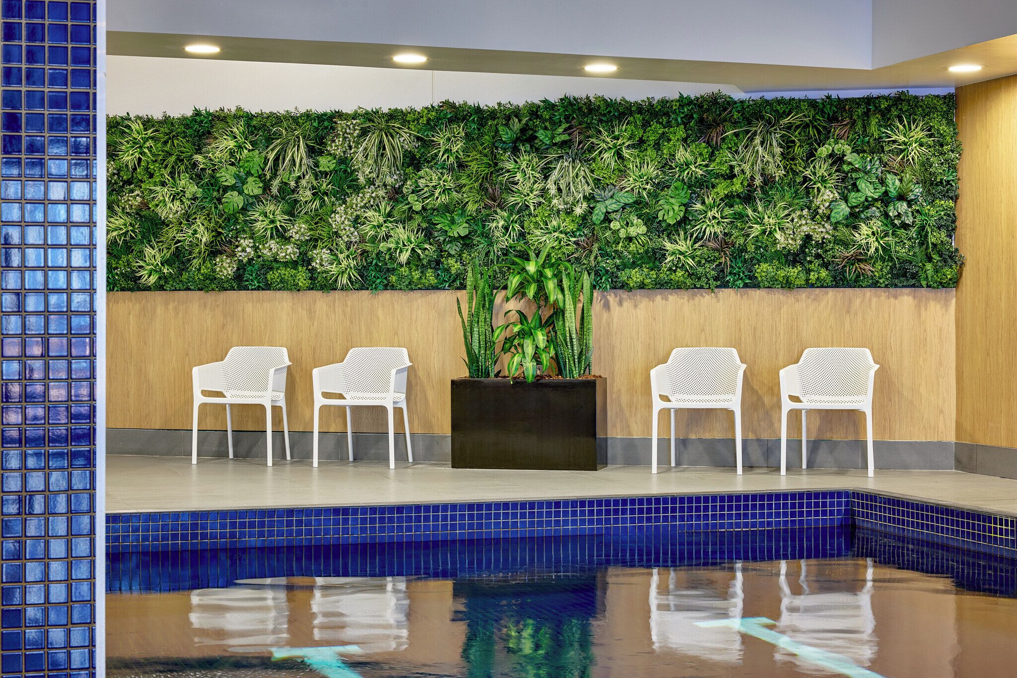 Uma área de piscina com piso cerâmico, parede verde com plantas e cadeiras brancas.