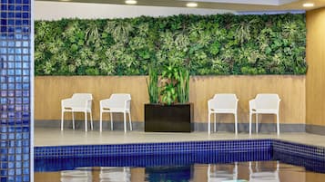 Uma área de piscina com piso cerâmico, parede verde com plantas e cadeiras brancas.