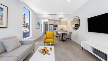 Uma sala de estar moderna com sofá, uma mesa de centro com comida, uma poltrona amarela, área de refeições com cadeiras, cozinha com eletrodomésticos e uma televisão grande.