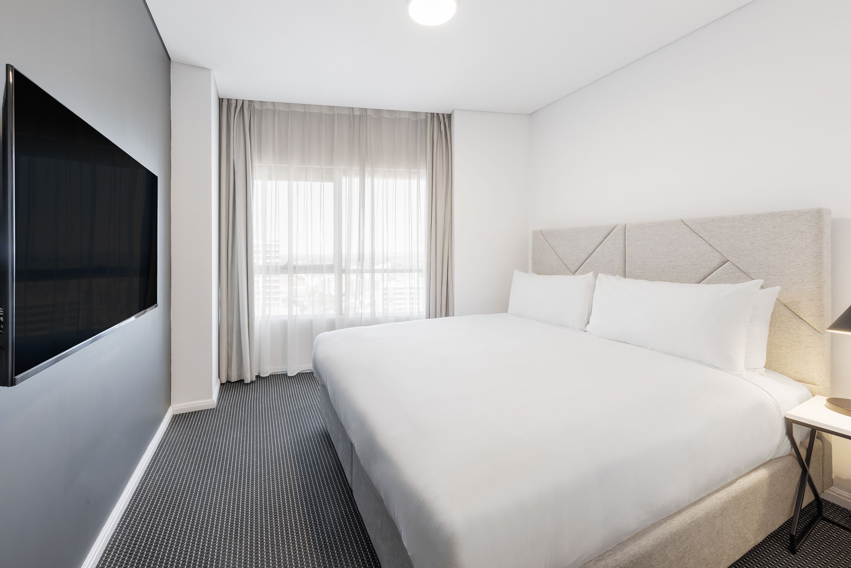 Quarto de hotel com uma cama grande, televisão de tela plana na parede, janela com cortinas leves e uma mesinha de cabeceira com abajur.