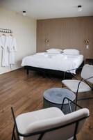 Quarto de hotel moderno com uma cama grande, duas mesas de cabeceira, uma cadeira e um porta-robes fixado na parede.