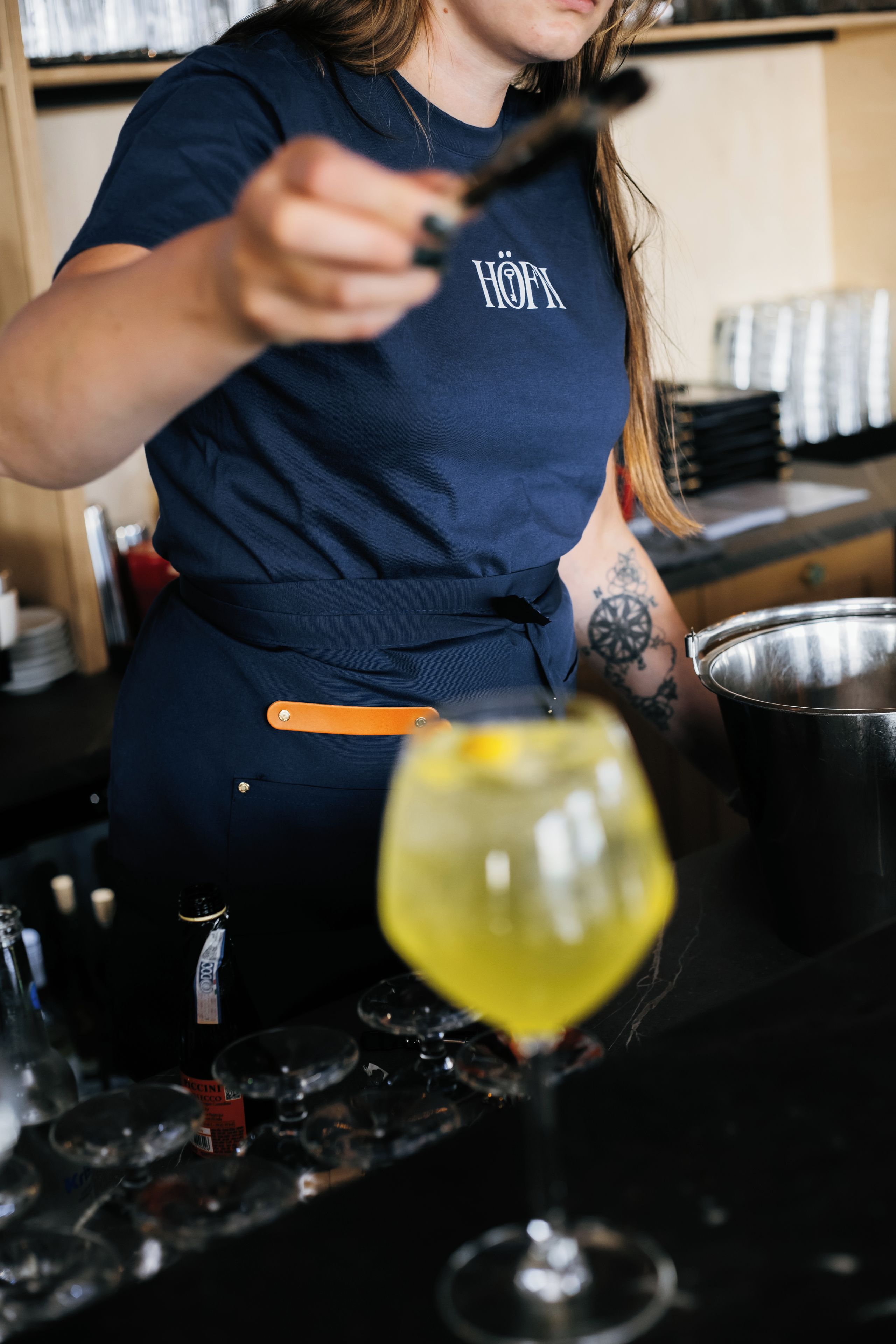 Uma pessoa vestindo camisa azul escura com um logotipo, preparando uma bebida com líquido amarelo num copo.