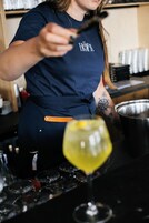 Uma pessoa vestindo camisa azul escura com um logotipo, preparando uma bebida com líquido amarelo num copo.