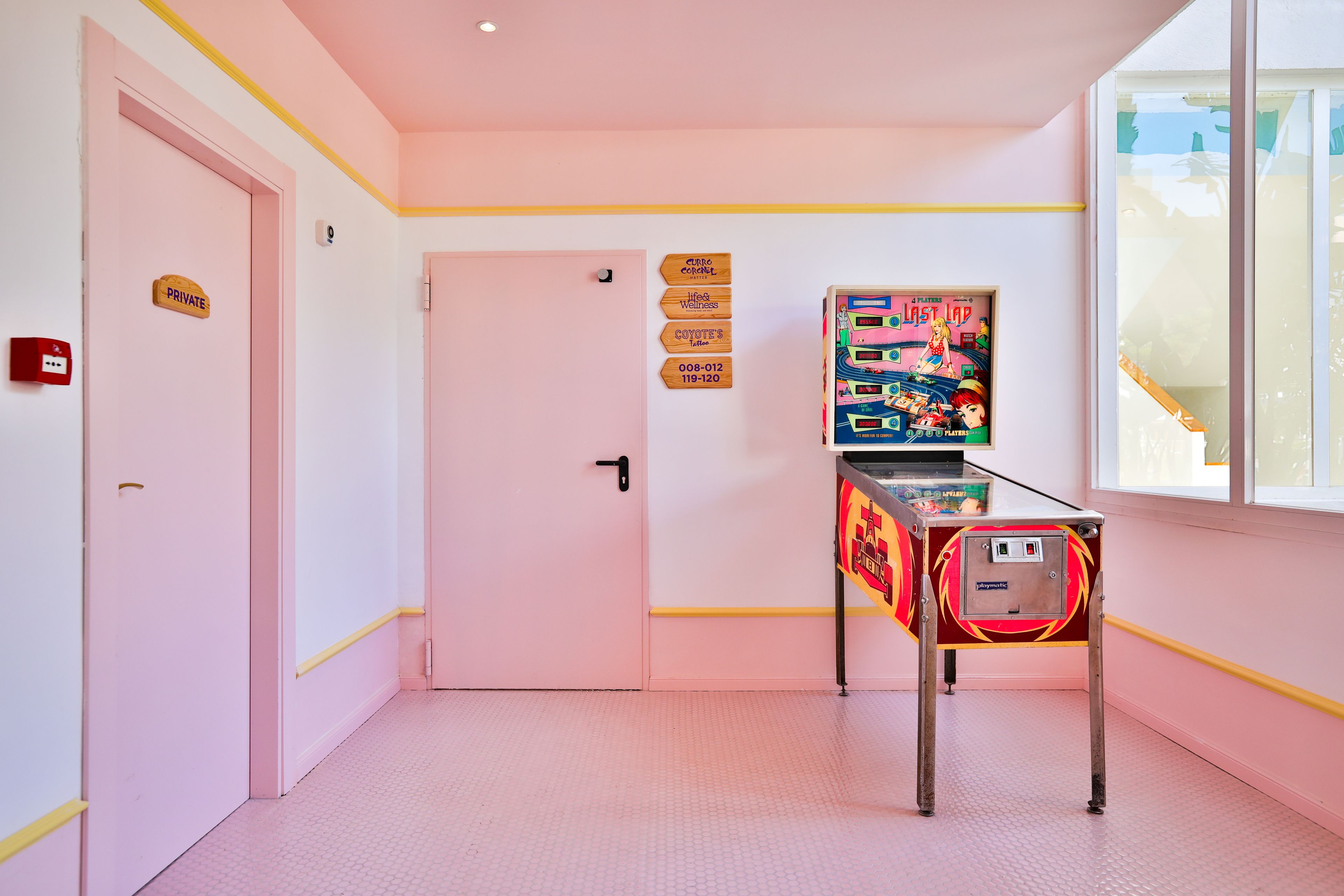 Um corredor com porta privativa, alarme de incêndio e uma mesa com pôster colorido.