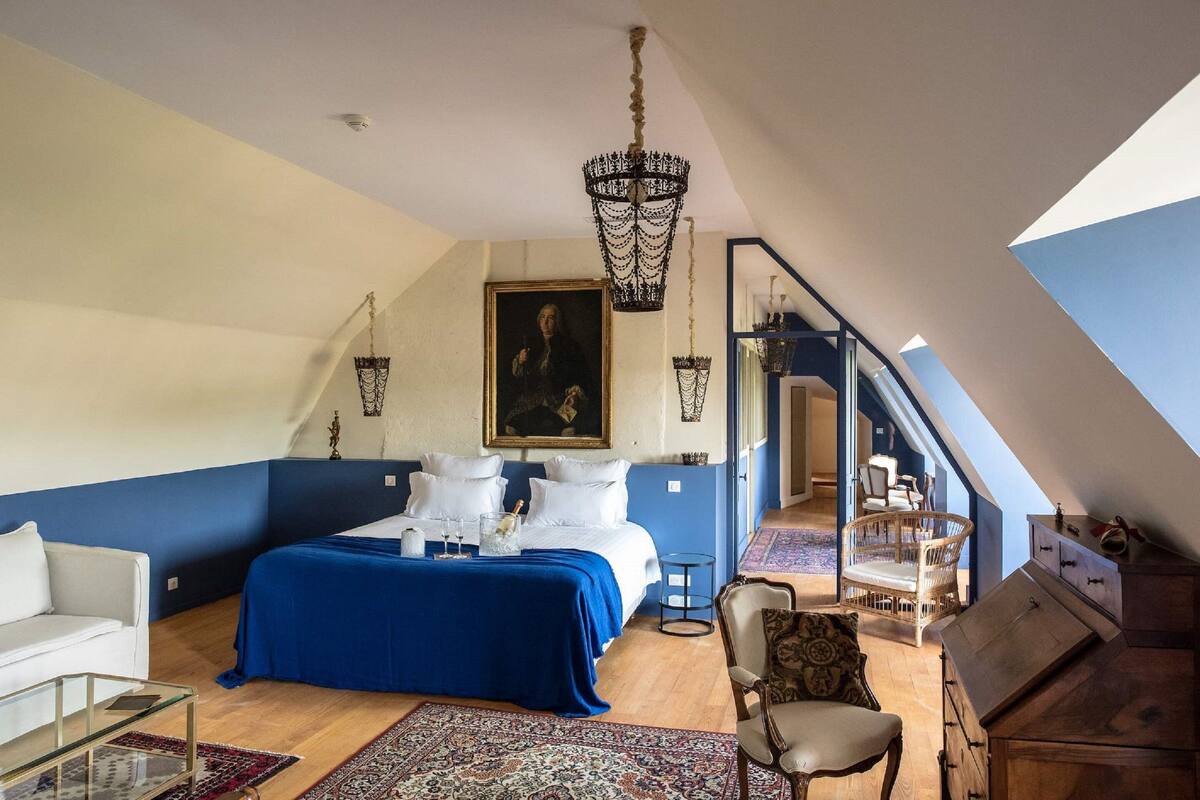 Quarto com cama grande, lustre, pintura e escada para outro espaço.