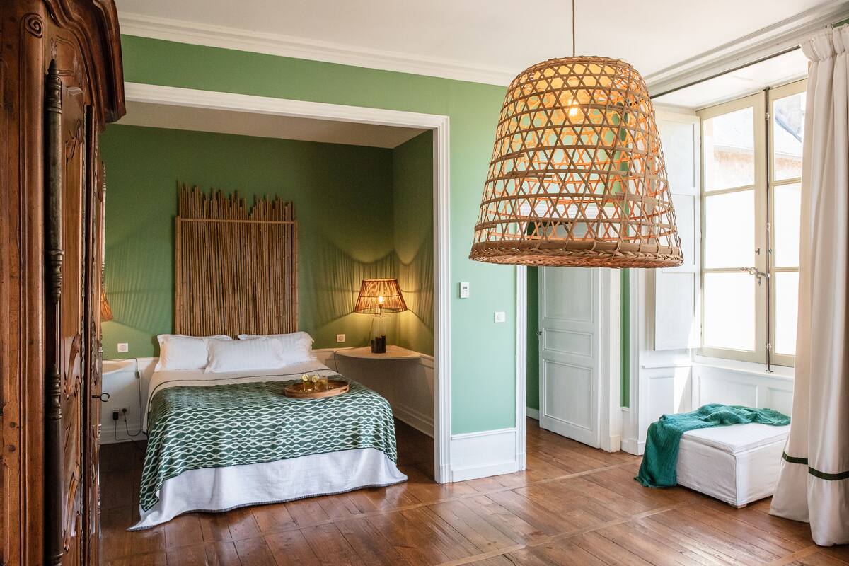 Quarto com cama, mesa de cabeceira, luminária e janela com cortinas.