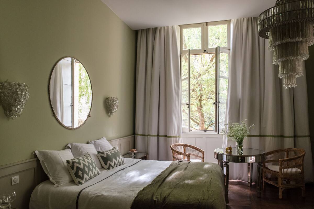 Quarto com cama, espelho redondo, janela com cortinas, mesa com um vaso e duas cadeiras de vime.