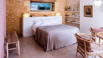 Um quarto com parede de pedra, uma cama com manta cinza, uma cadeira de vime, uma mesa pequena e prateleiras com plantas e um quadro.