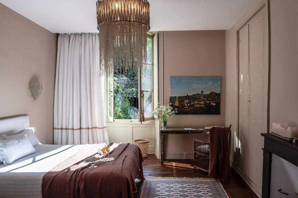 Quarto com cama, escrivaninha, cadeira, janela com cortinas, pintura e lustre.