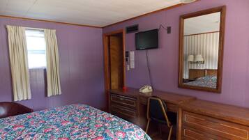 Quarto com uma cama, escrivaninha, cadeira, cômoda, televisão e espelho.