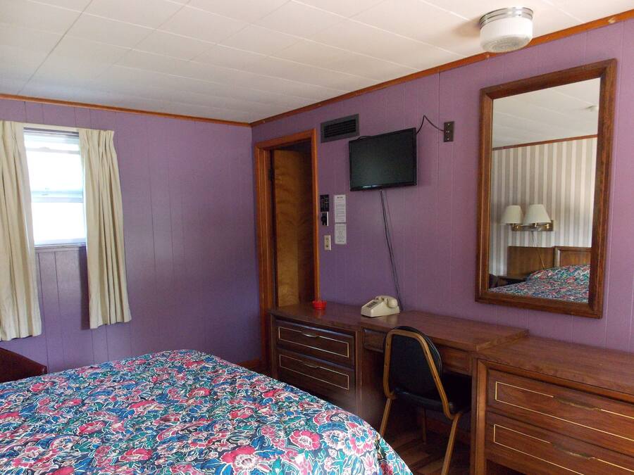 Quarto com uma cama, escrivaninha, cadeira, cômoda, televisão e espelho.