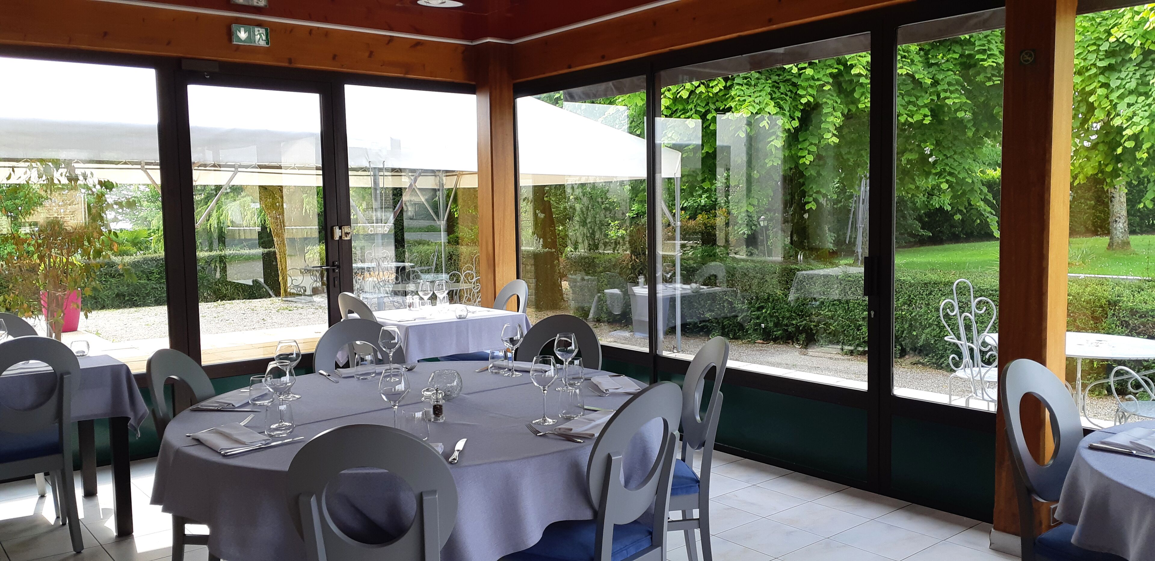 Restaurante com mesas redondas preparadas para refeições, grandes janelas com vista para o verde e teto de madeira.