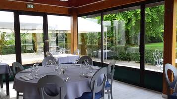 Restaurante com mesas redondas preparadas para refeições, grandes janelas com vista para o verde e teto de madeira.