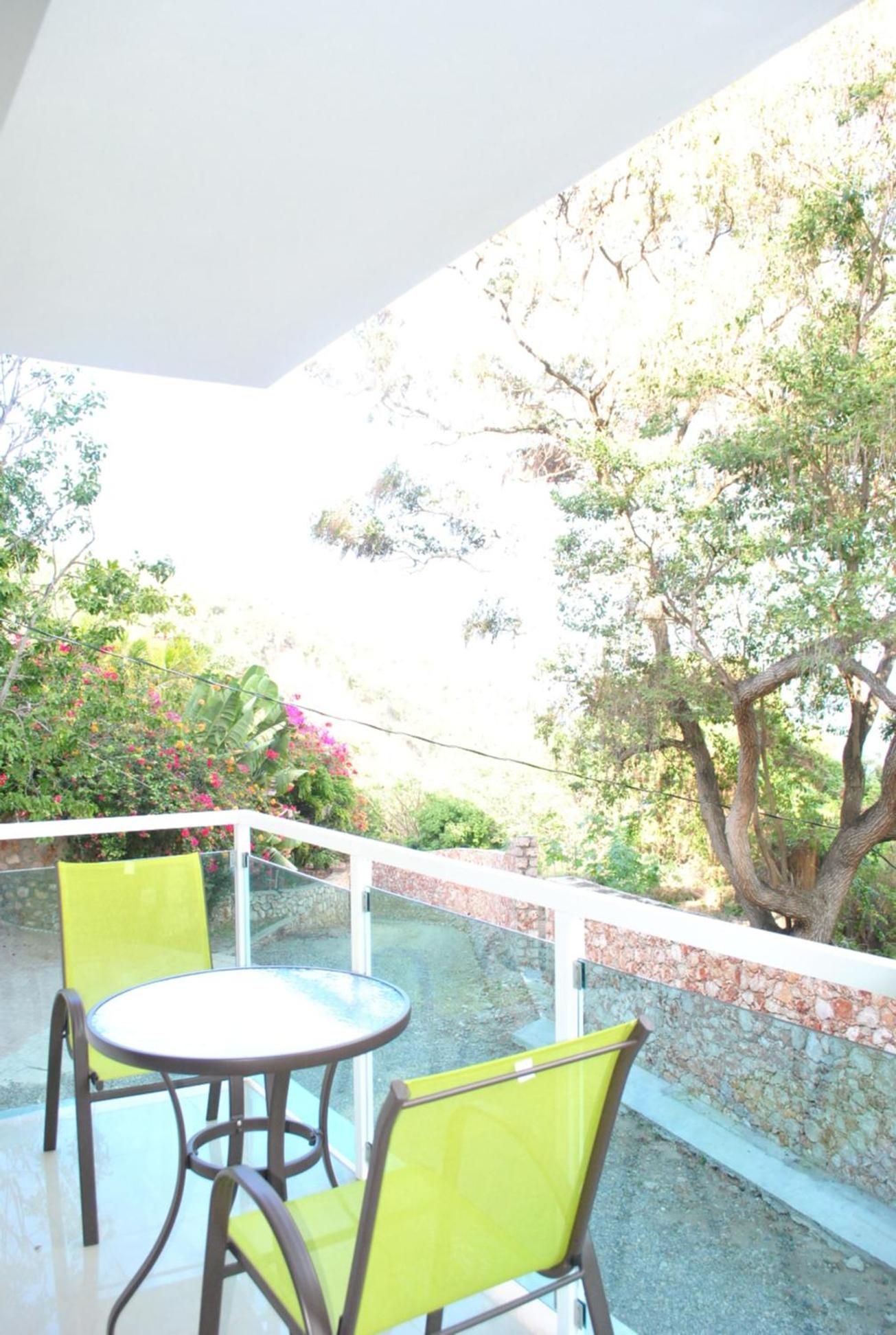 Uma varanda com uma mesa redonda e duas cadeiras, com vista para um jardim com árvores e arbustos.