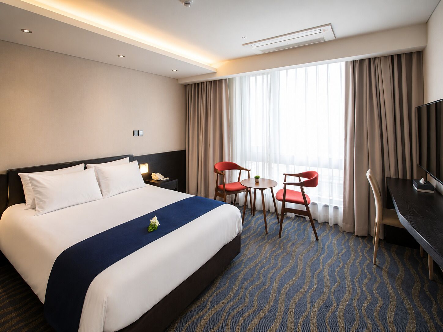 Quarto de hotel com uma cama, escrivaninha, cadeira, mesinha e televisão.