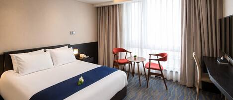 Quarto de hotel com uma cama, escrivaninha, cadeira, mesinha e televisão.