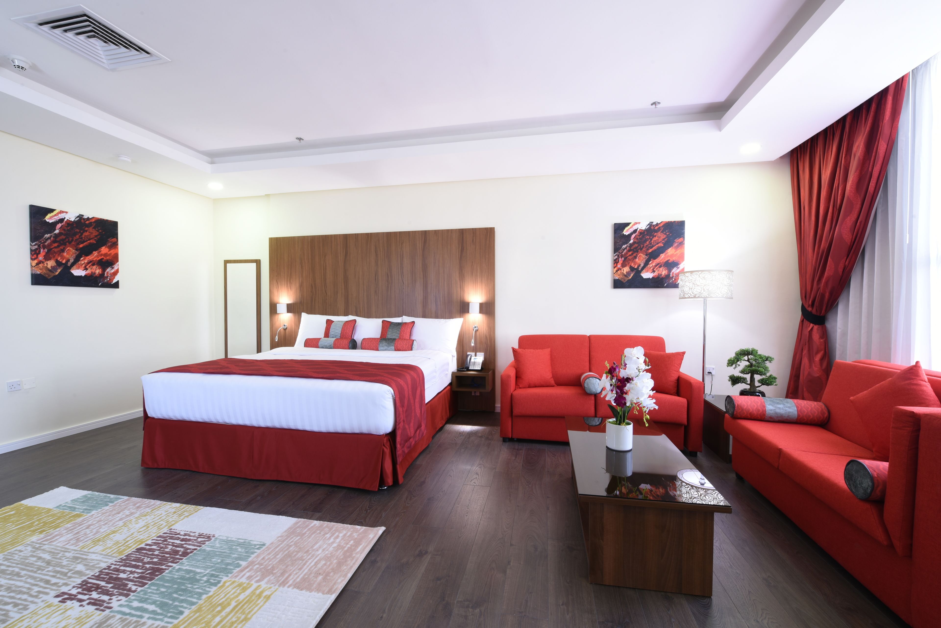 Quarto de hotel com uma cama grande, um sofá vermelho, uma mesa de centro e duas luminárias fixadas na parede.