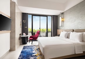 Quarto de hotel moderno com uma cama grande, televisão, uma pequena mesa com cadeira e vista para o verde através de grandes janelas.
