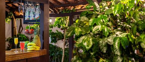 Uma área de bar com balcão de madeira, taças de vinho penduradas e frutas tropicais como bananas e abacaxi.