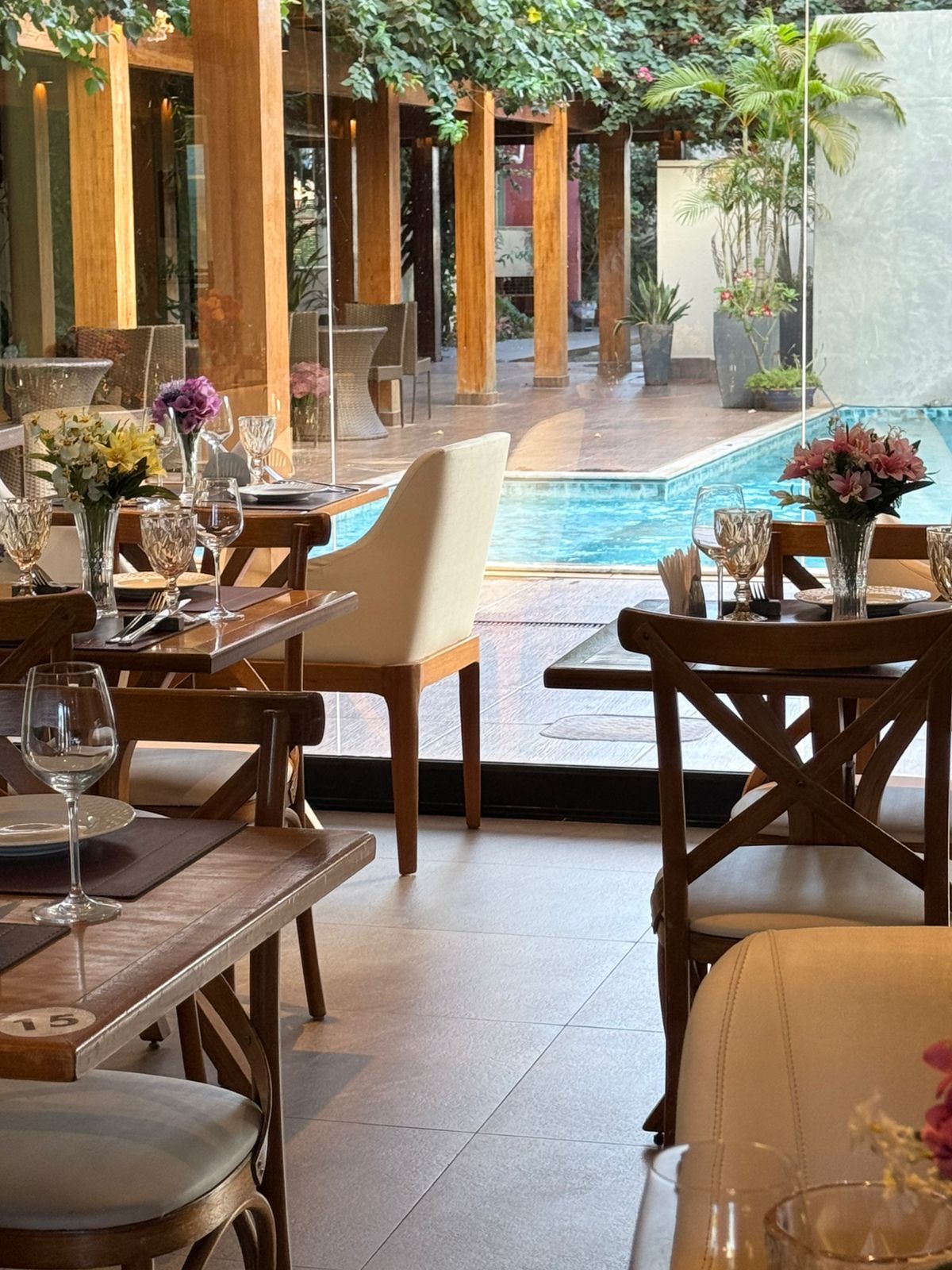 Restaurante com área externa e mesas próximas a uma piscina, com mesas e cadeiras de madeira e plantas em vasos.