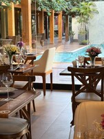 Restaurante com área externa e mesas próximas a uma piscina, com mesas e cadeiras de madeira e plantas em vasos.