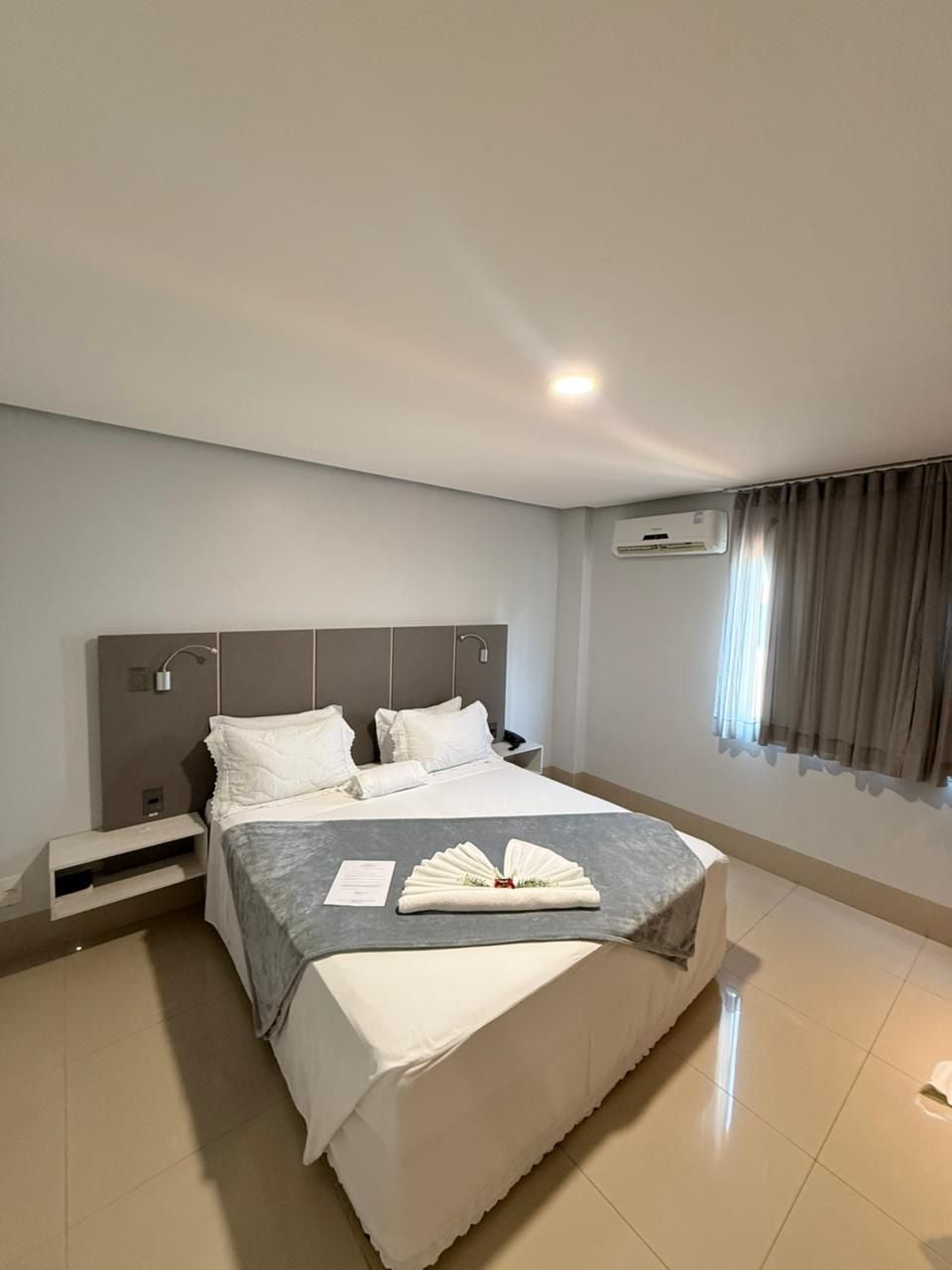 Quarto de hotel com cama grande, mesa de cabeceira, ar condicionado e janela com cortinas.