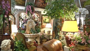 Um quarto com uma fonte, uma estátua, uma cadeira, uma mesa com um abajur e diversas plantas em vasos.