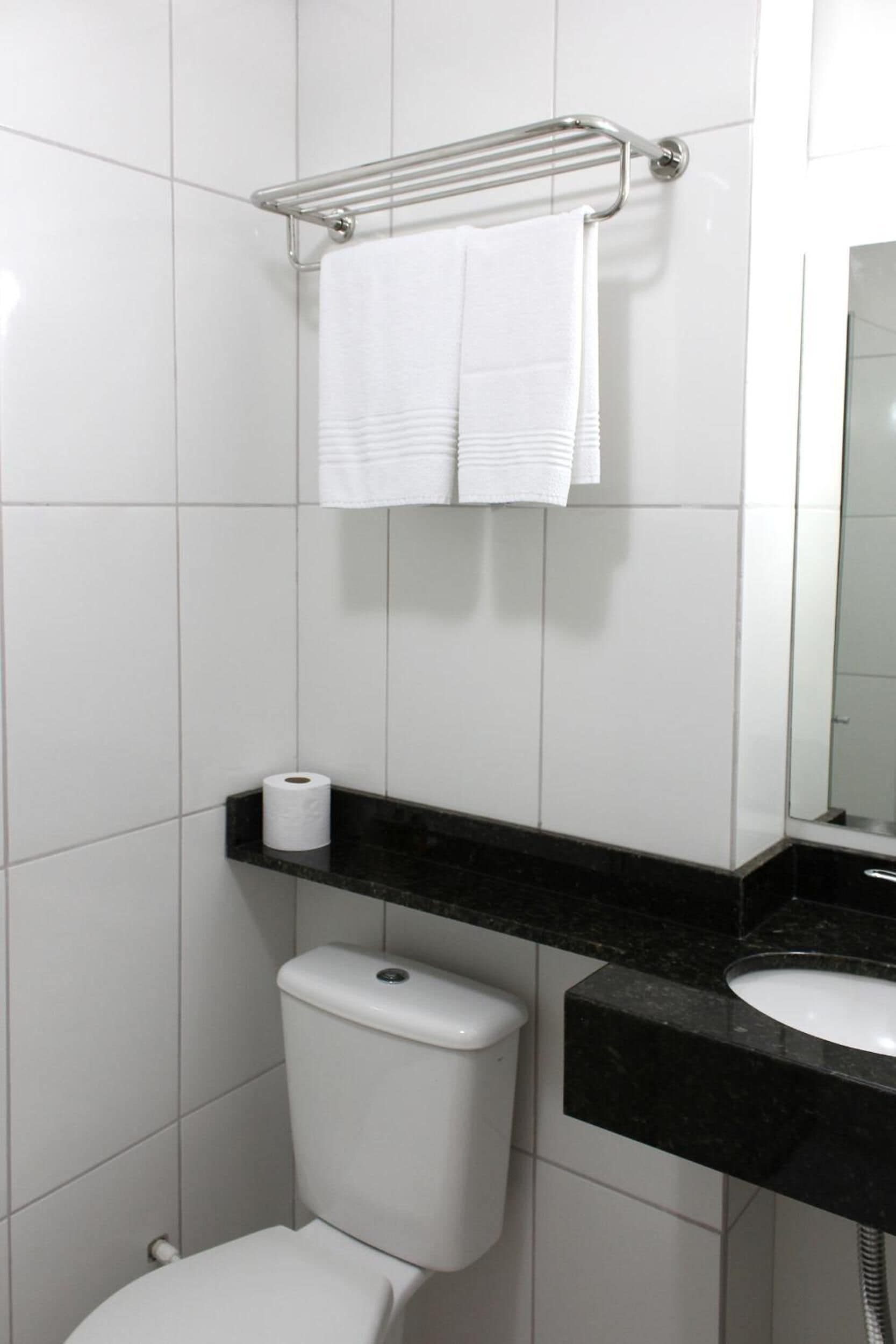 Um banheiro com vaso sanitário, pia, porta-toalhas com duas toalhas e rolo de papel higiênico em uma prateleira.