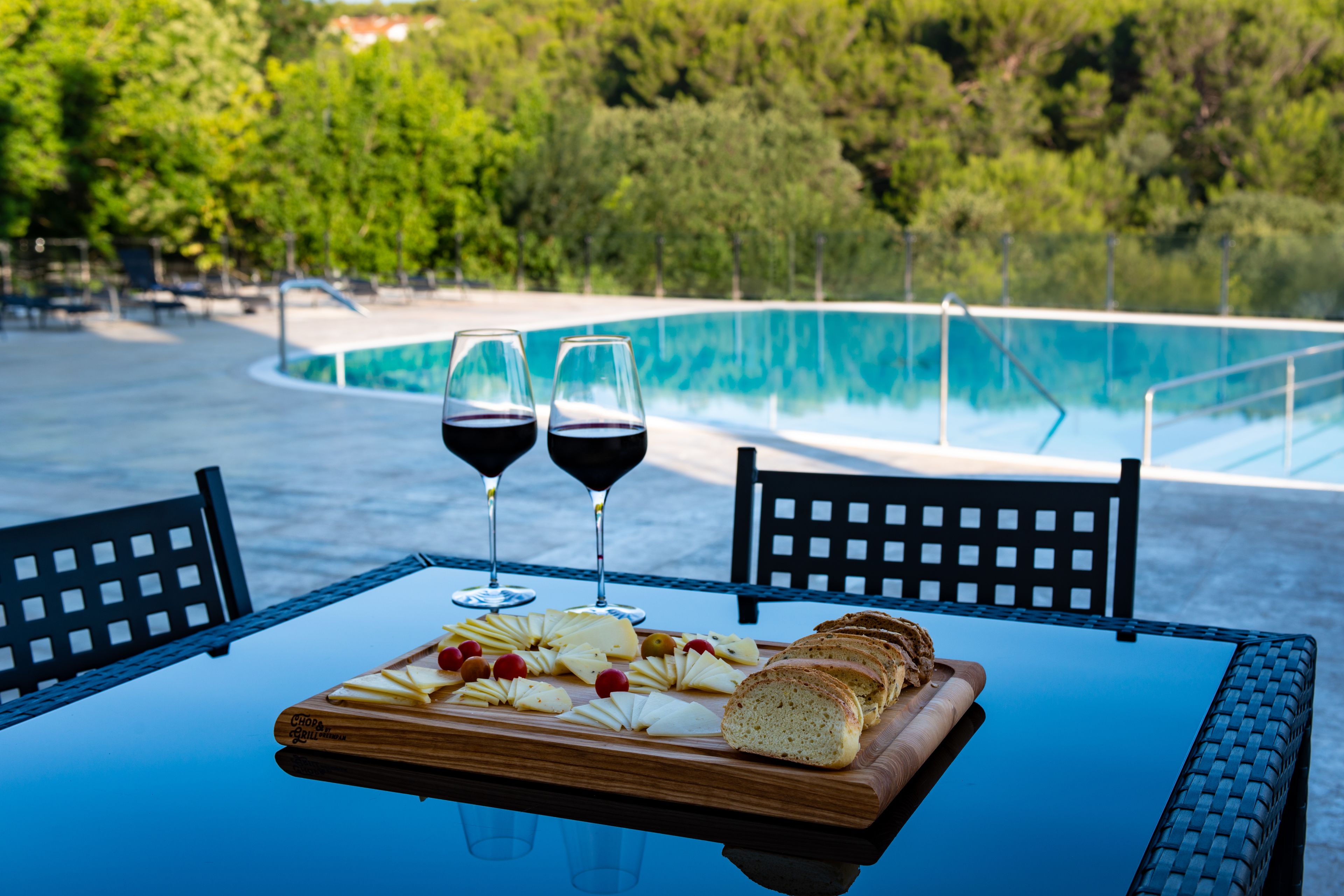 Uma mesa com uma tábua de queijos, dois copos de vinho tinto e vista para uma piscina e vegetação.