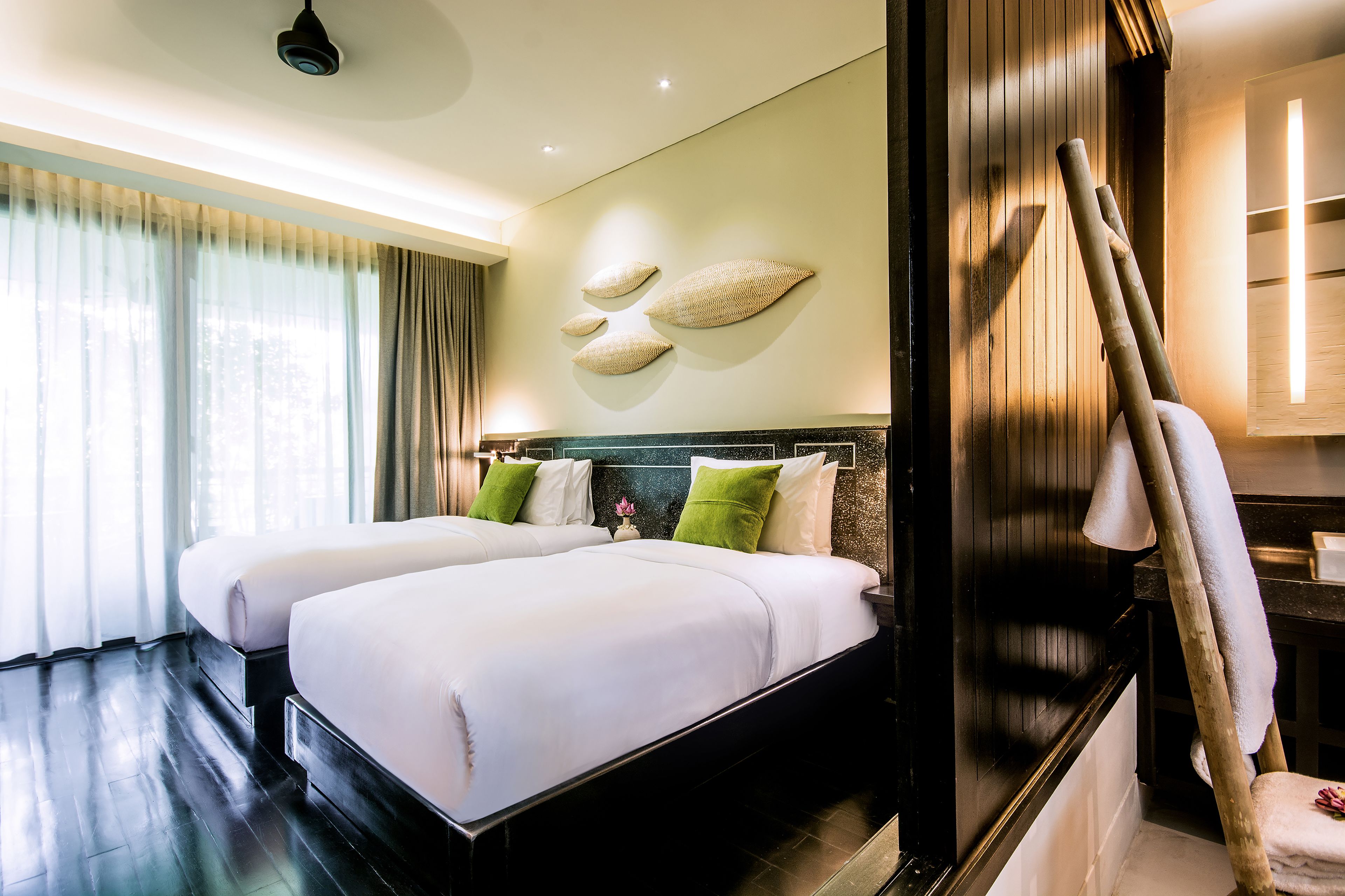 Quarto de hotel com duas camas, uma janela grande com cortinas, um objeto decorativo na parede e um porta-toalhas de madeira.