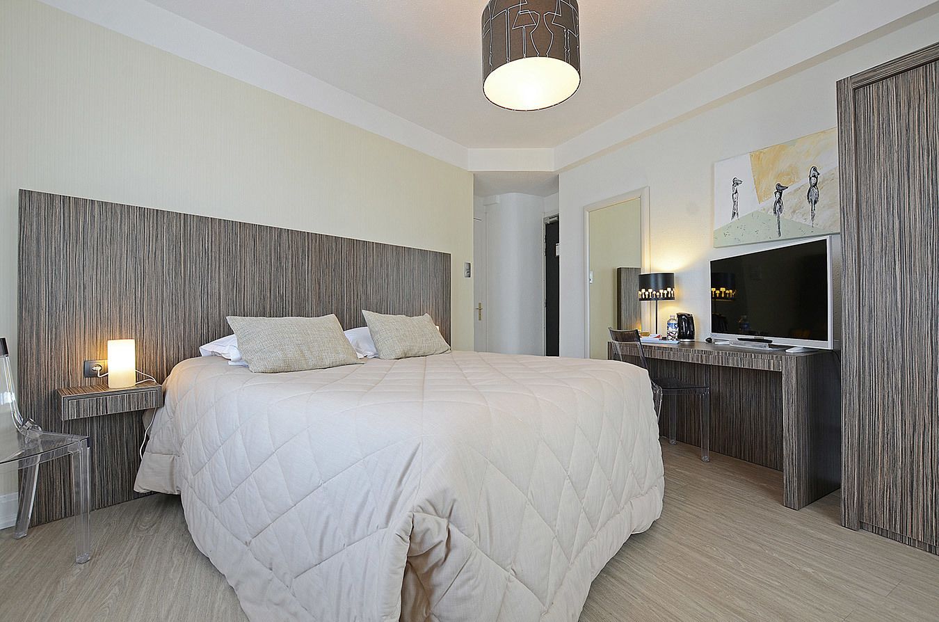 Quarto de hotel com uma cama grande, uma televisão, uma escrivaninha com abajur e um quadro na parede.