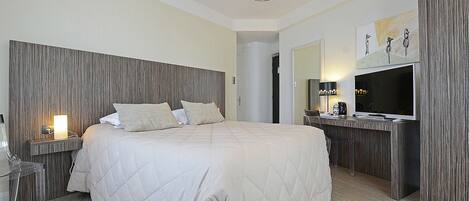 Quarto de hotel com uma cama grande, uma televisão, uma escrivaninha com abajur e um quadro na parede.