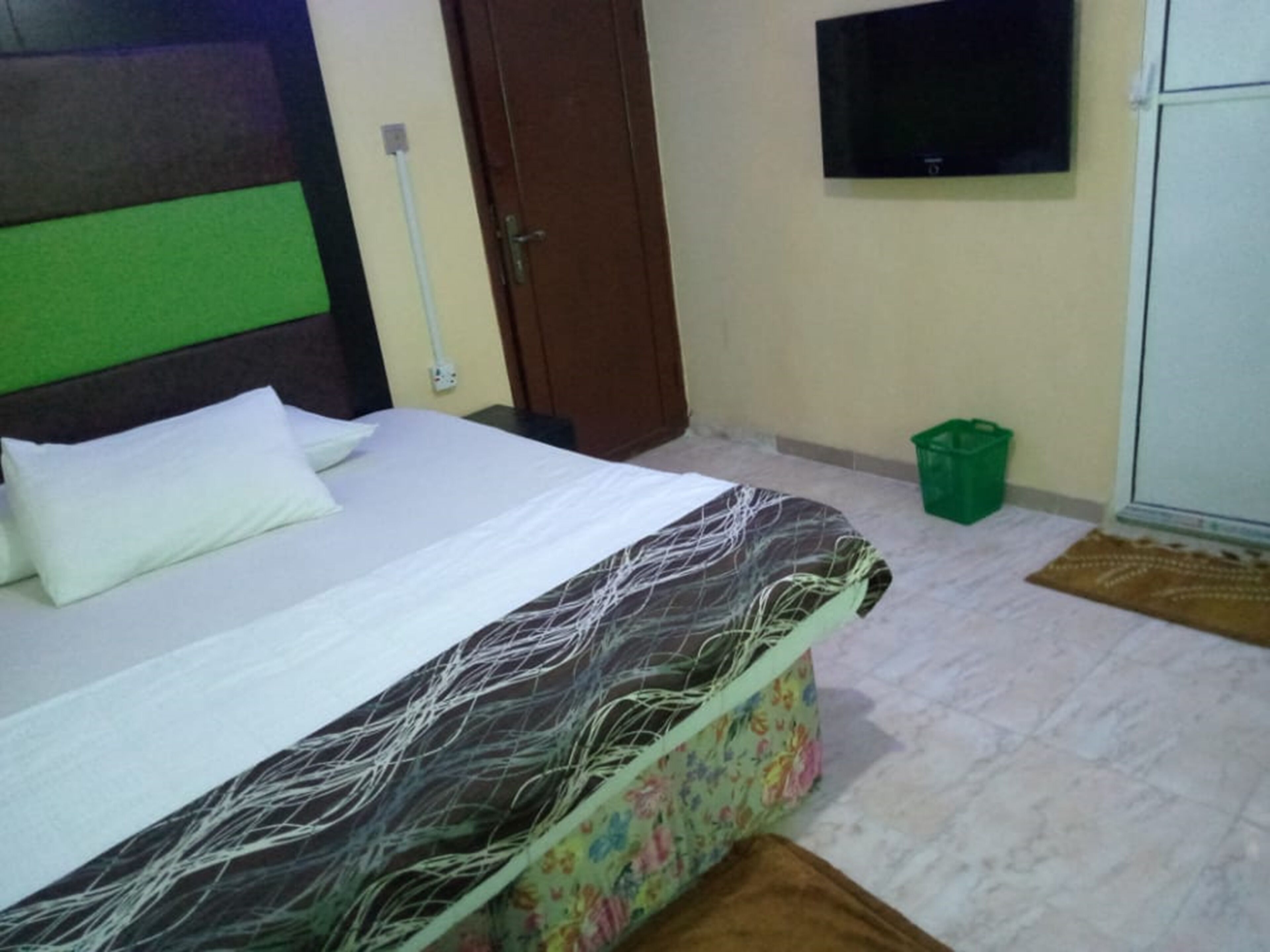 Quarto com cama, televisão na parede, porta e lixeira.