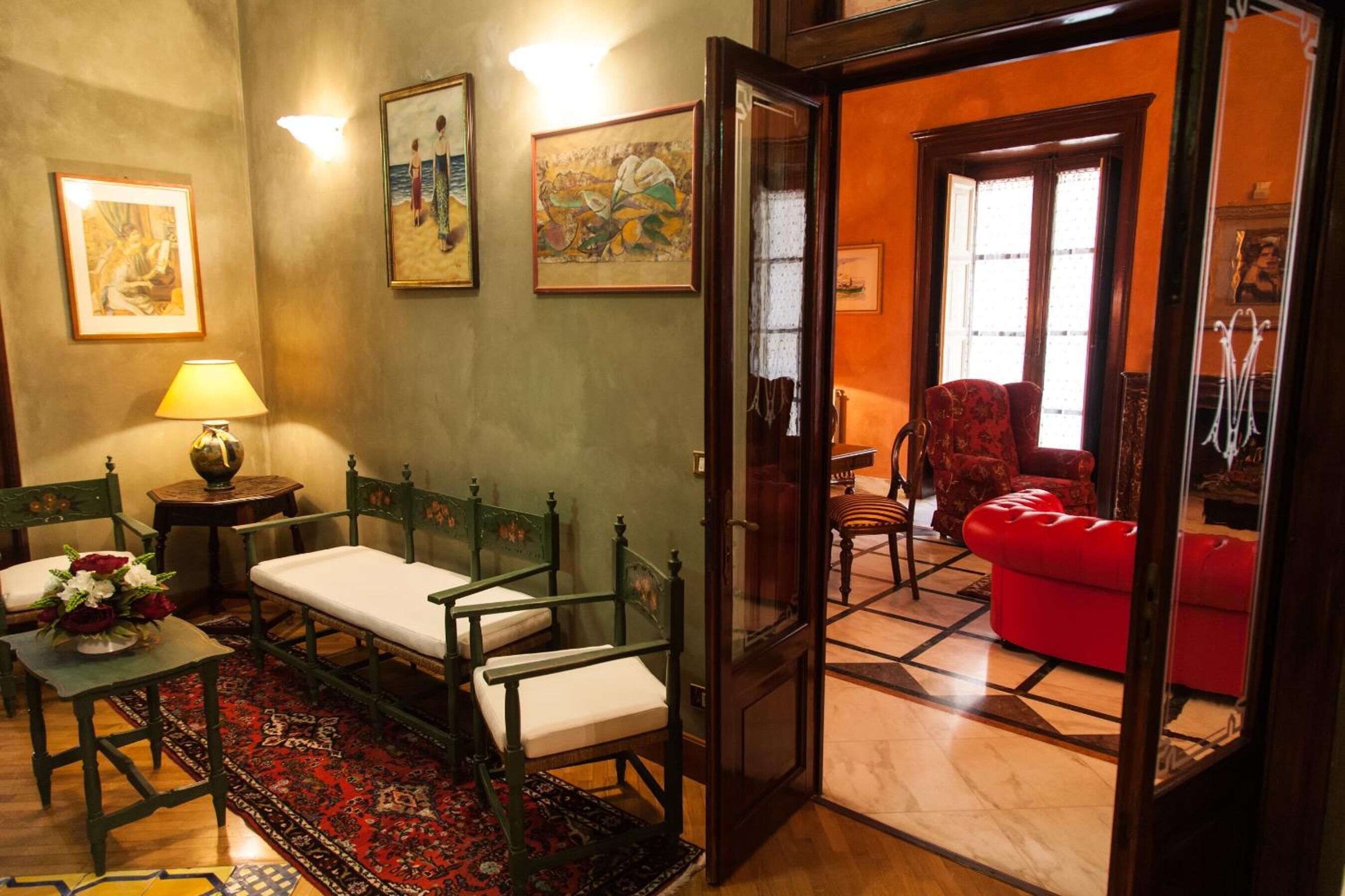Um quarto com piso de madeira, um sofá vermelho, uma poltrona verde, um banco branco e um tapete estampado.