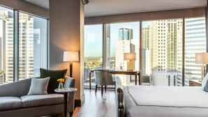 Quarto de hotel com uma cama grande, um sofá, uma pequena mesa com abajur e uma cadeira. Janelas amplas oferecem vista para a cidade.