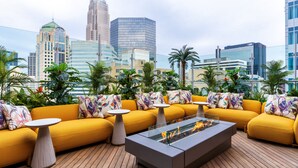 Um lounge no terraço com sofás amarelos, uma mesa de centro de vidro e um elemento decorativo com fogo.