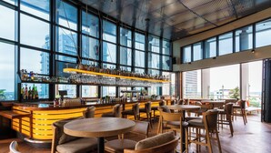 Uma área de bar moderna com teto alto, janelas amplas e diversas opções de assento, incluindo mesas redondas e cadeiras de vime.