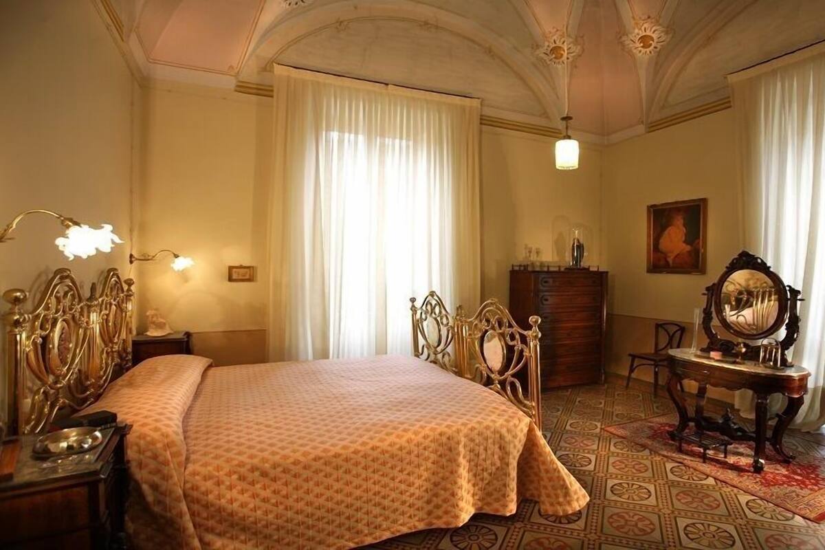 Quarto de hotel com uma cama grande, cômoda de madeira, espelho e lustre.