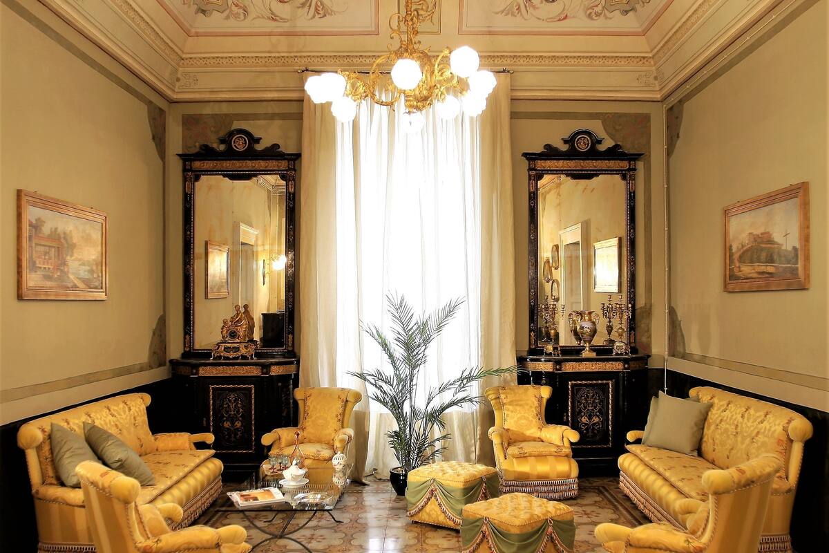 Quarto luxuoso com decoração ornamental no teto, um candelabro e duas grandes espelhos. Há quatro poltronas confortáveis e uma mesa de centro com objetos decorativos.