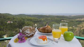 Uma mesa com croissants, uma maçã verde, um copo de suco de laranja e um prato com sobremesa, com vista para uma paisagem de montanhas.
