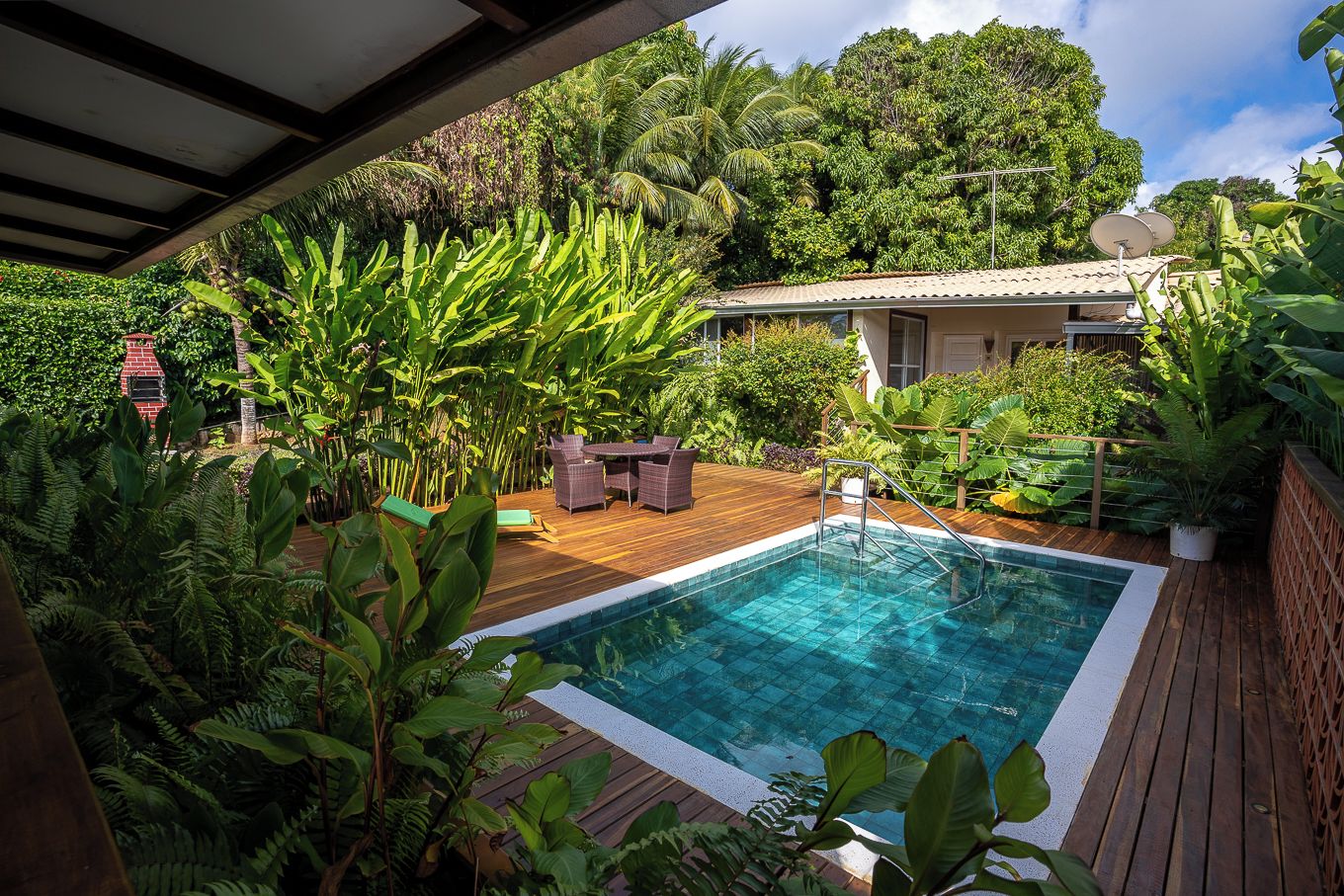 Área de piscina com deck de madeira, rodeada por vegetação exuberante e uma casa ao fundo.