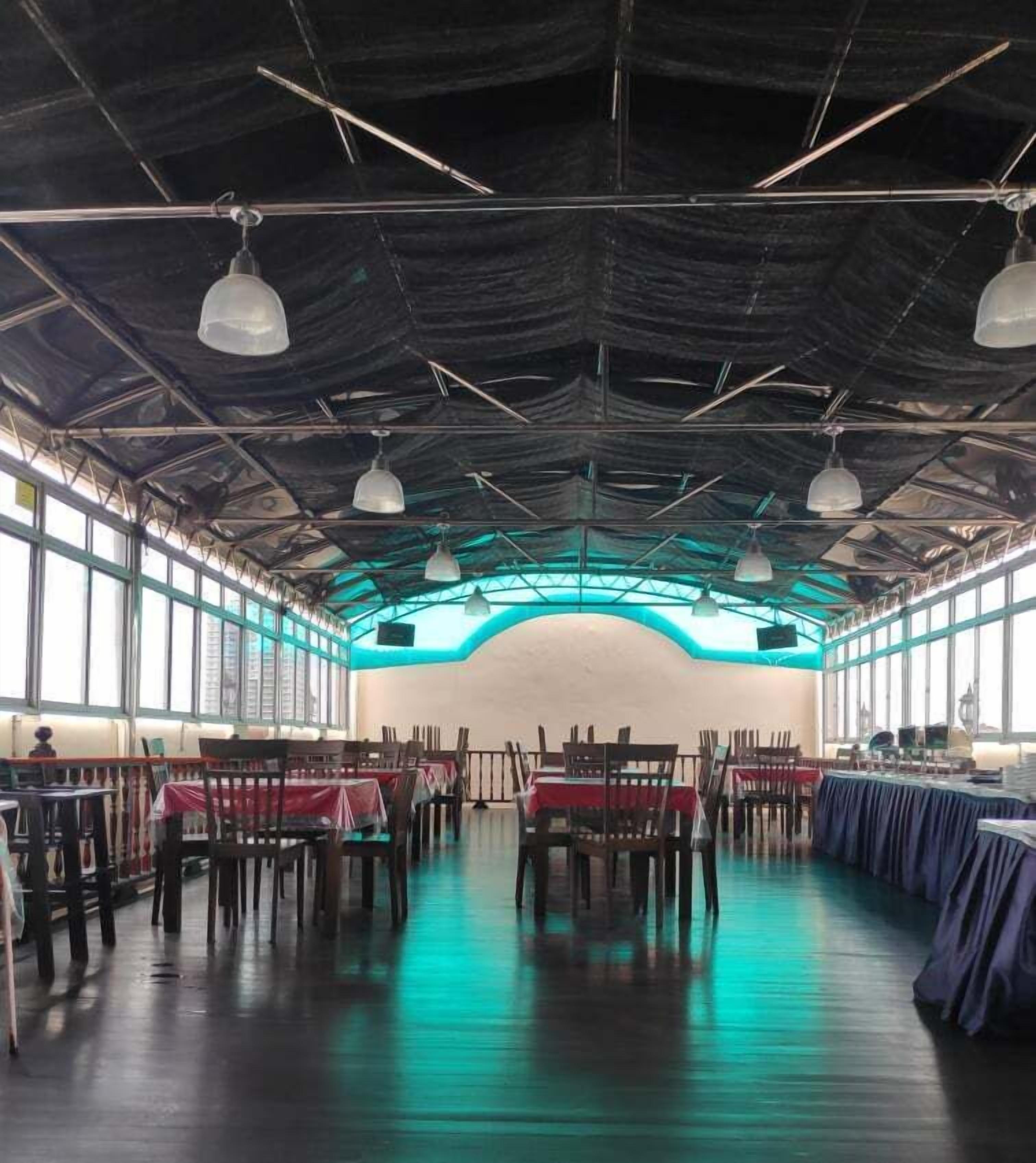 Um espaço interno com mesas e cadeiras, teto alto com vigas aparentes e amplas janelas que permitem entrada de luz natural.