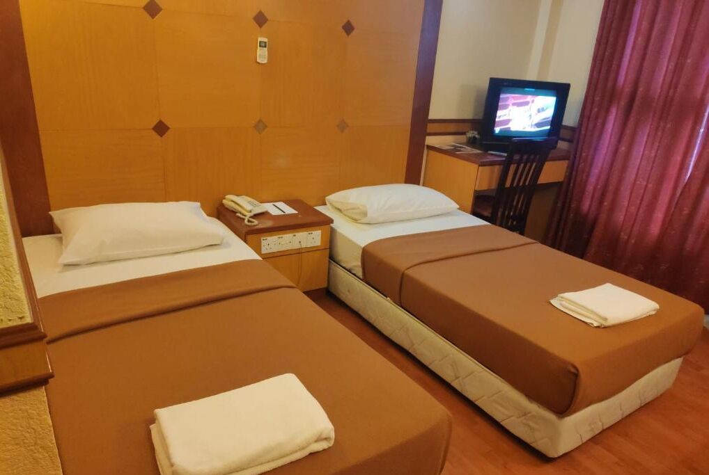 Quarto de hotel com duas camas de solteiro, uma mesa de trabalho com televisão e uma cadeira.