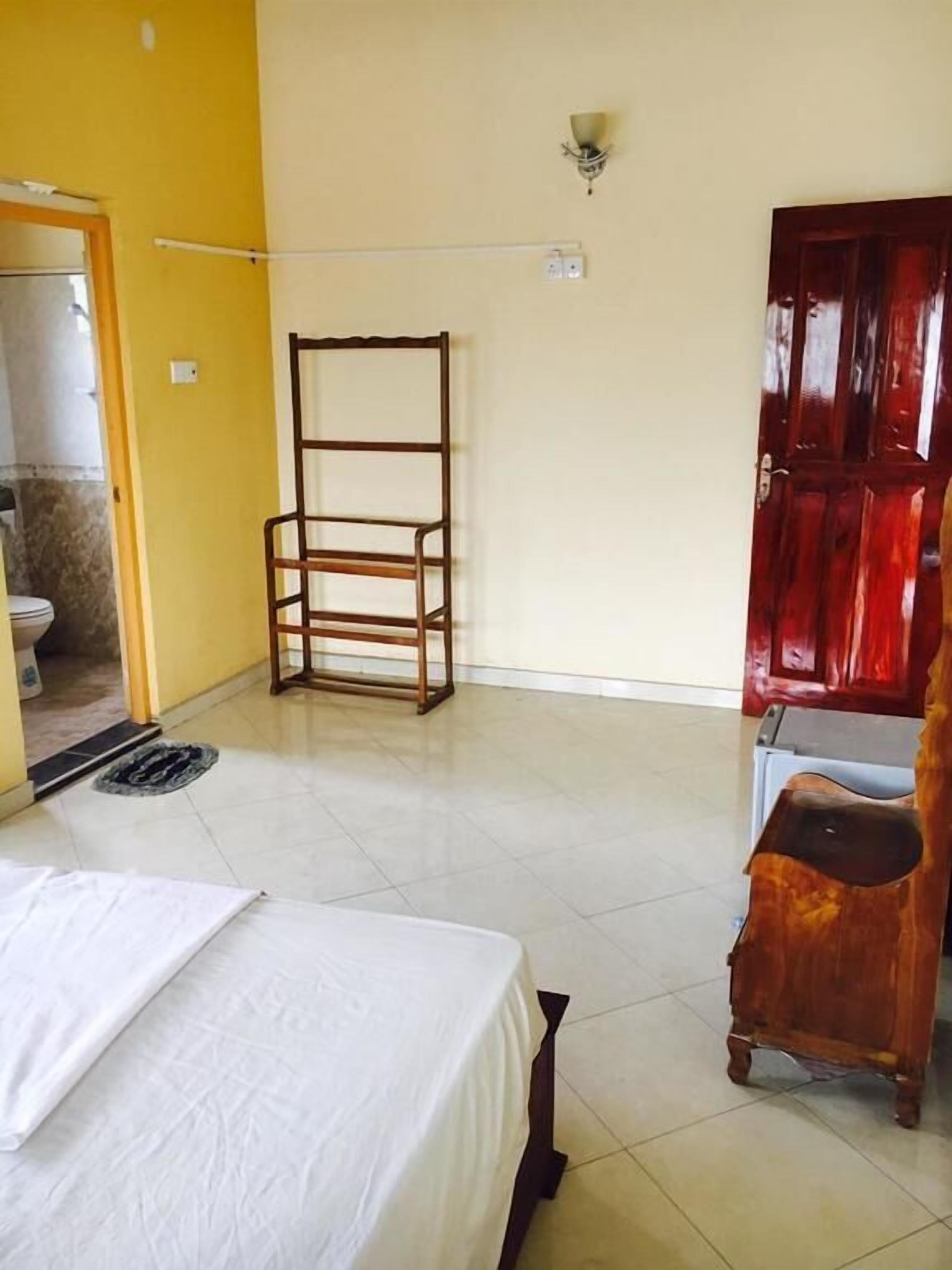 Um quarto com uma cama, uma prateleira de madeira, uma porta vermelha e um aparelho de iluminação na parede.