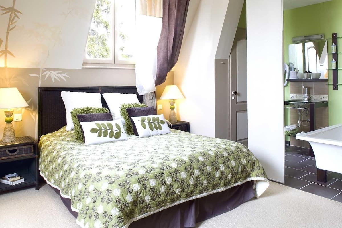 Quarto com cama, duas mesas de cabeceira, um criado-mudo, janela com cortinas, banheiro com pia e espelho, e parede verde.