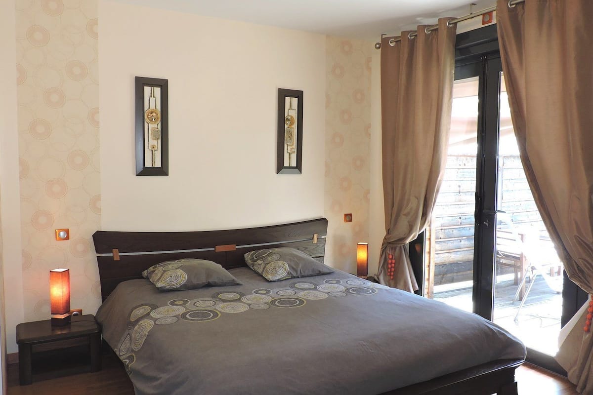 Quarto com uma cama grande, dois quadros pendurados na parede, uma mesa de cabeceira com abajur e vista para o exterior através de uma janela com cortinas.
